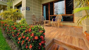 Um edifício moderno com fachada em azulejos, um deck de madeira com mobiliário exterior e um jardim bem cuidado com flores vermelhas.