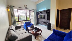 Uma sala de estar com sofá, uma poltrona azul, uma mesa de centro com um laptop, uma TV fixada na parede e um ventilador de teto.