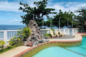 Piscina com borda curva, cercada por um muro branco, com cadeiras e uma formação rochosa.