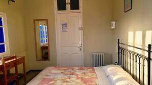 Quarto com cama, uma cadeira de madeira, porta branca e janela com persianas azuis.