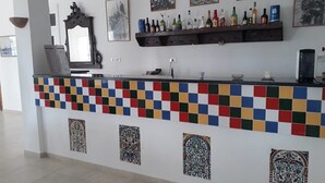 Uma área de bar com balcão revestido em azulejos, azulejos decorativos na parede, quadros emoldurados e uma prateleira com garrafas.