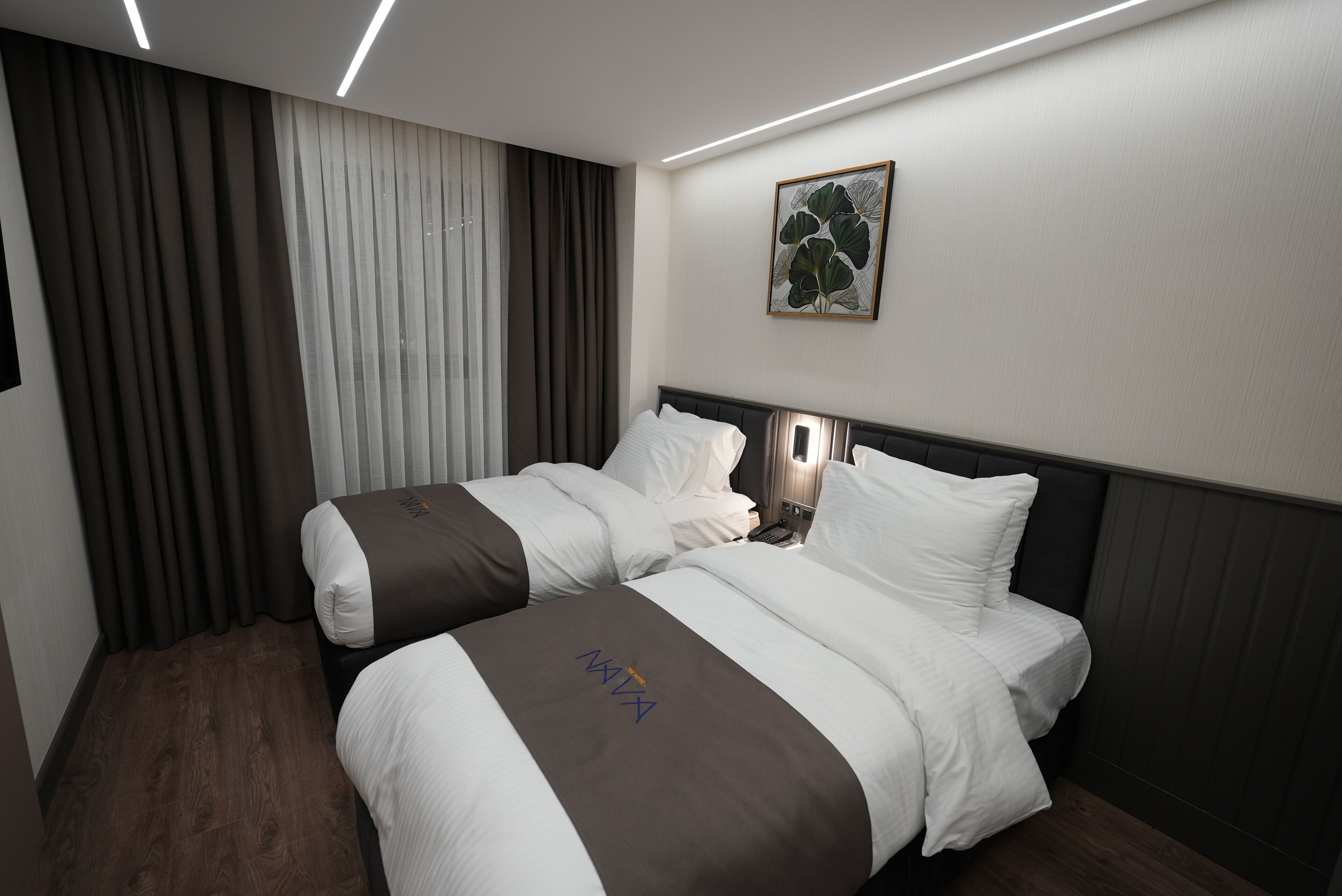 Quarto de hotel com duas camas, uma janela grande com cortinas, uma obra de arte na parede e uma luminária moderna.