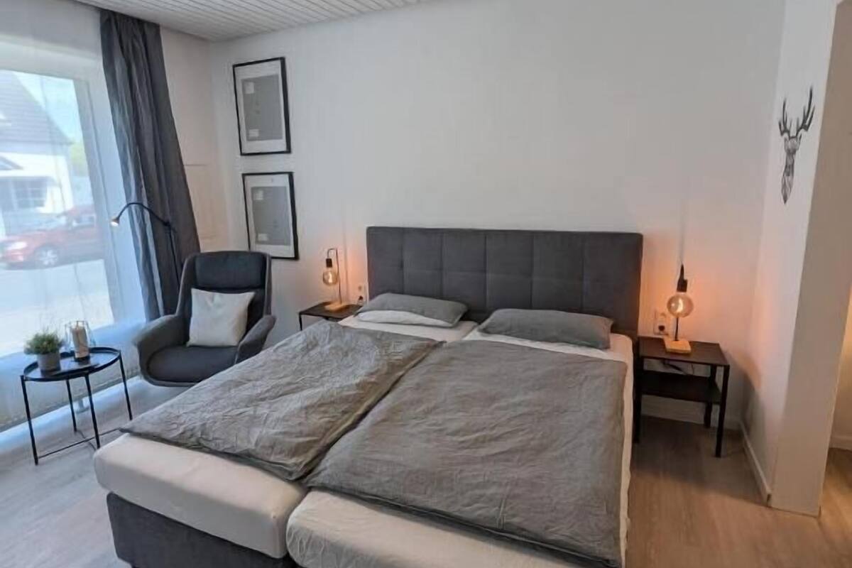 Quarto moderno com uma cama grande, uma cadeira, mesas de cabeceira e uma janela com cortinas.