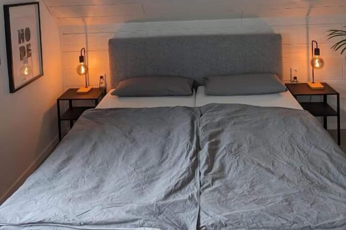 Quarto com cama beliche, duas mesas de cabeceira com luminárias, um quadro na parede e uma planta ao lado direito.