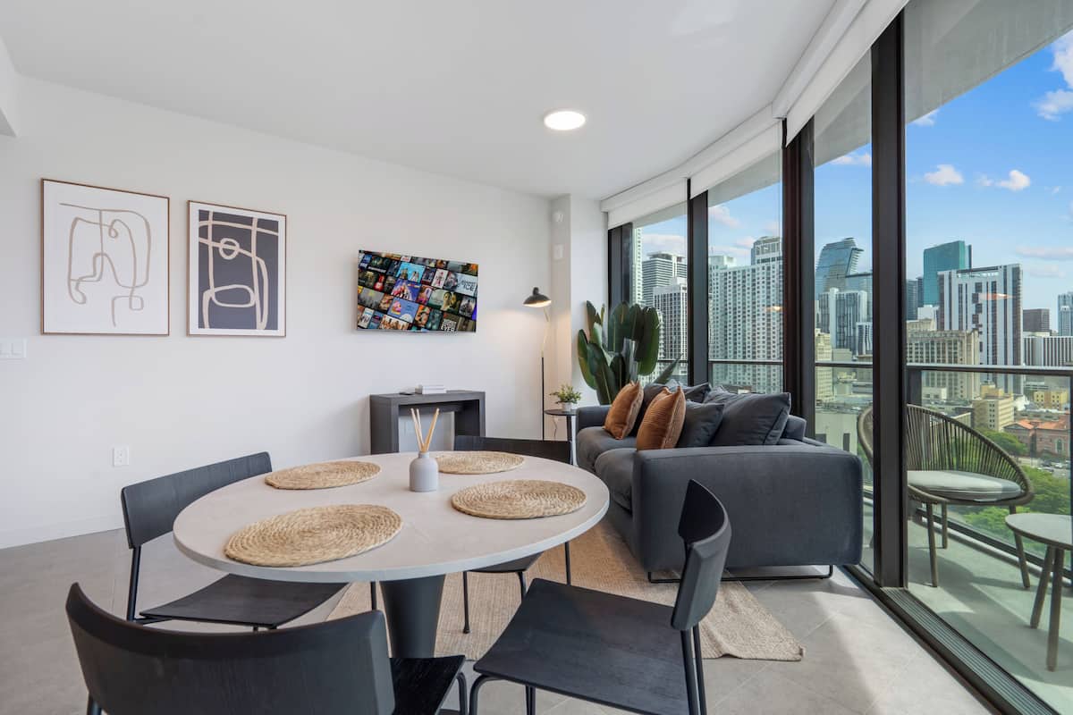 Sala de estar moderna com uma mesa redonda, cadeiras pretas, um sofá cinzento e vista para a cidade através de grandes janelas.