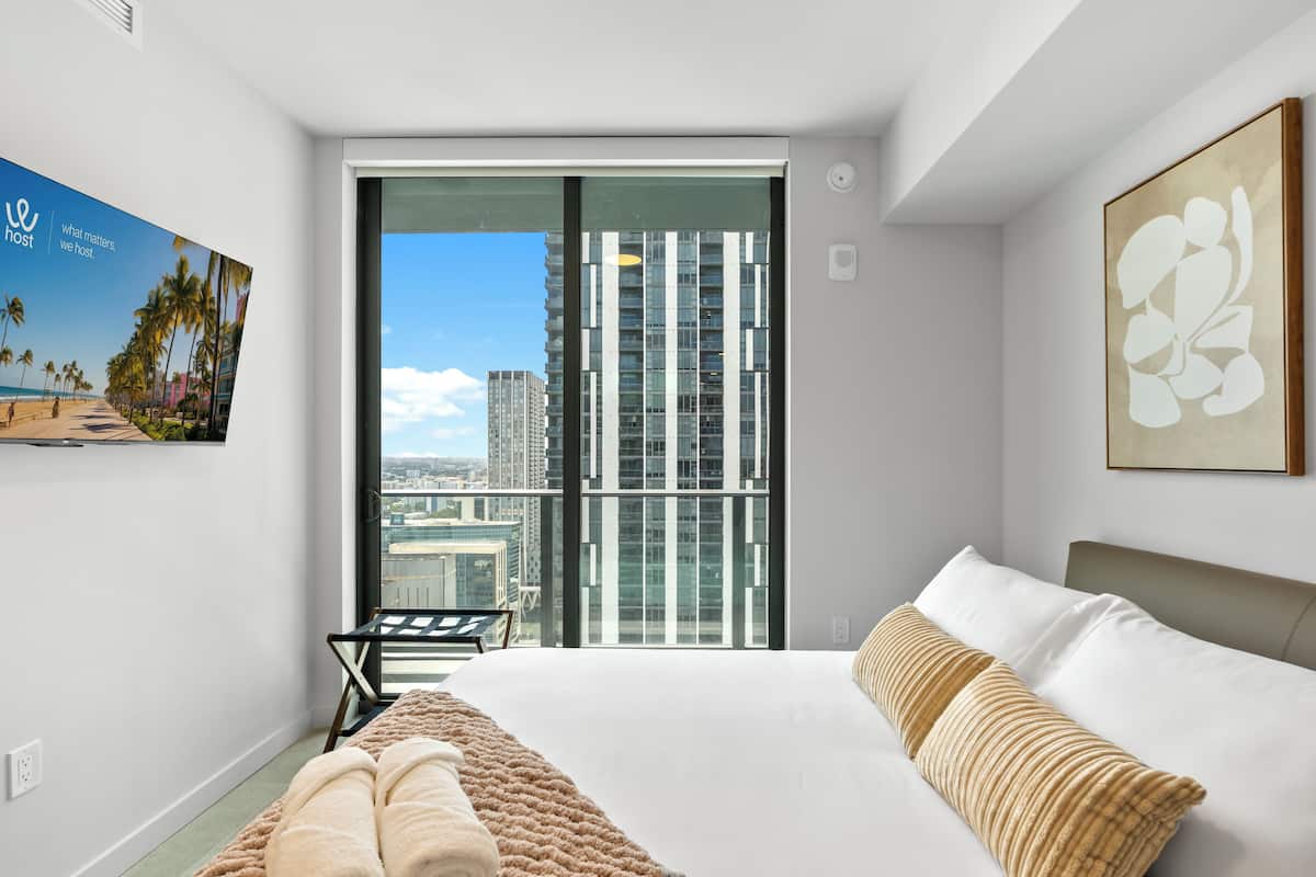 Quarto de hotel moderno com uma cama grande, vista para a cidade e uma varanda com porta de vidro.