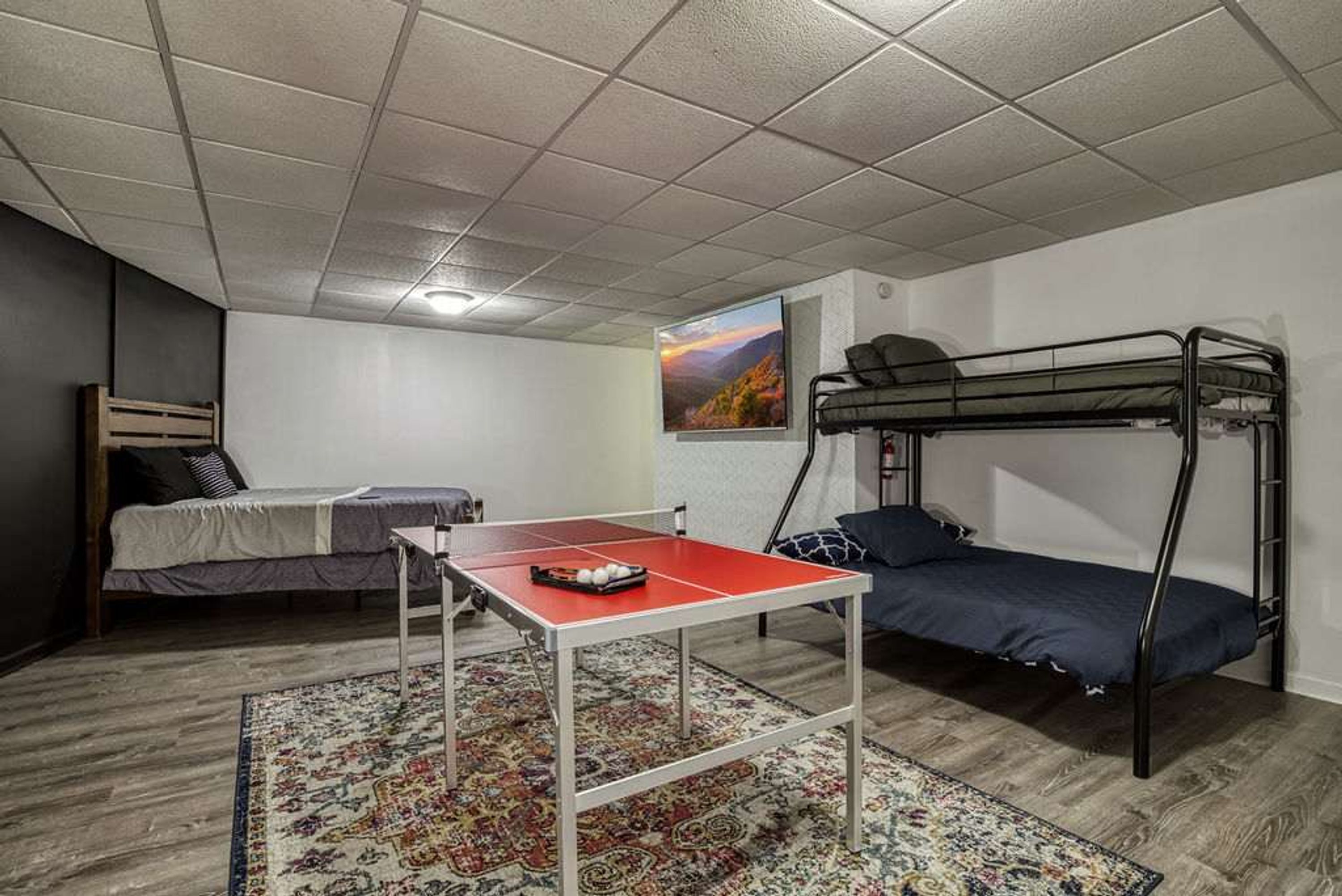 Quarto estilo dormitório com beliche, cama de solteiro, mesa pequena e tapete.