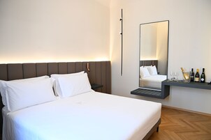 Quarto de hotel moderno com uma cama grande, mesas de cabeceira, um criado-mudo, espelho fixado na parede e uma estante com garrafas de champanhe.