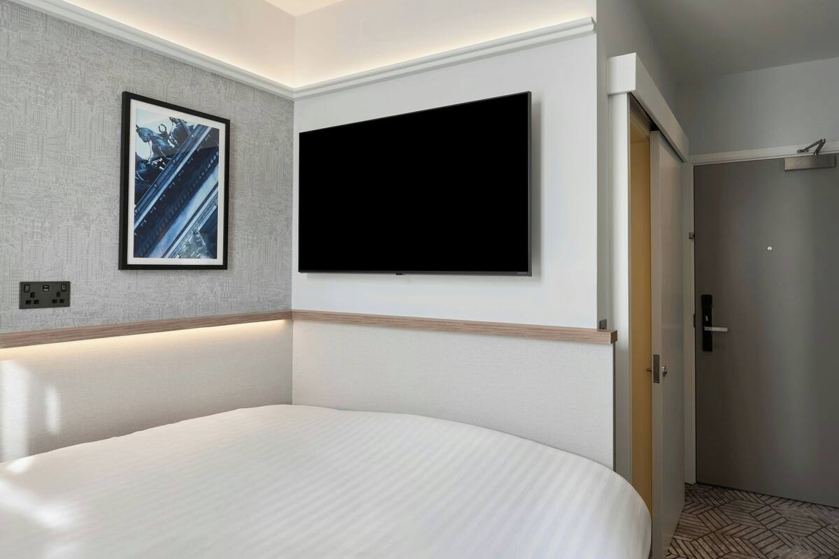 Quarto de hotel com uma cama, uma TV fixada na parede, um quadro emoldurado e uma porta que dá para outro cômodo.