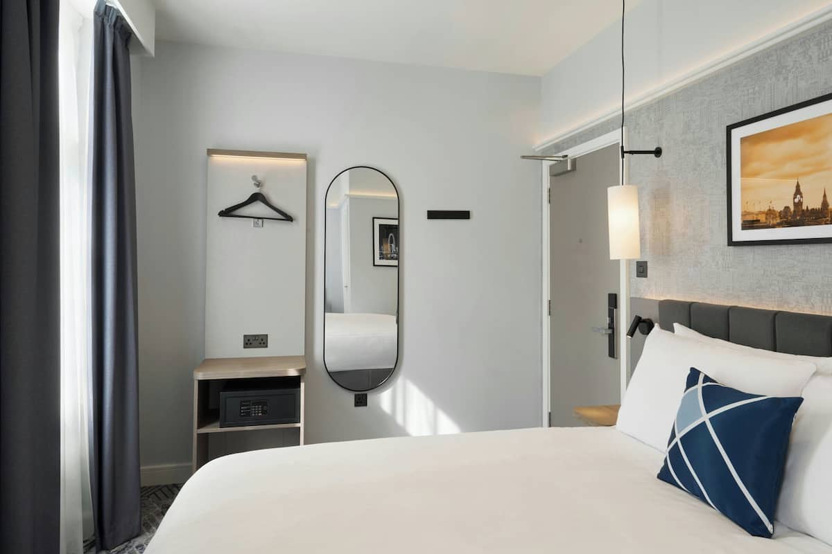Quarto de hotel com cama, mesa de cabeceira, espelho, cabideiro e um quadro na parede.