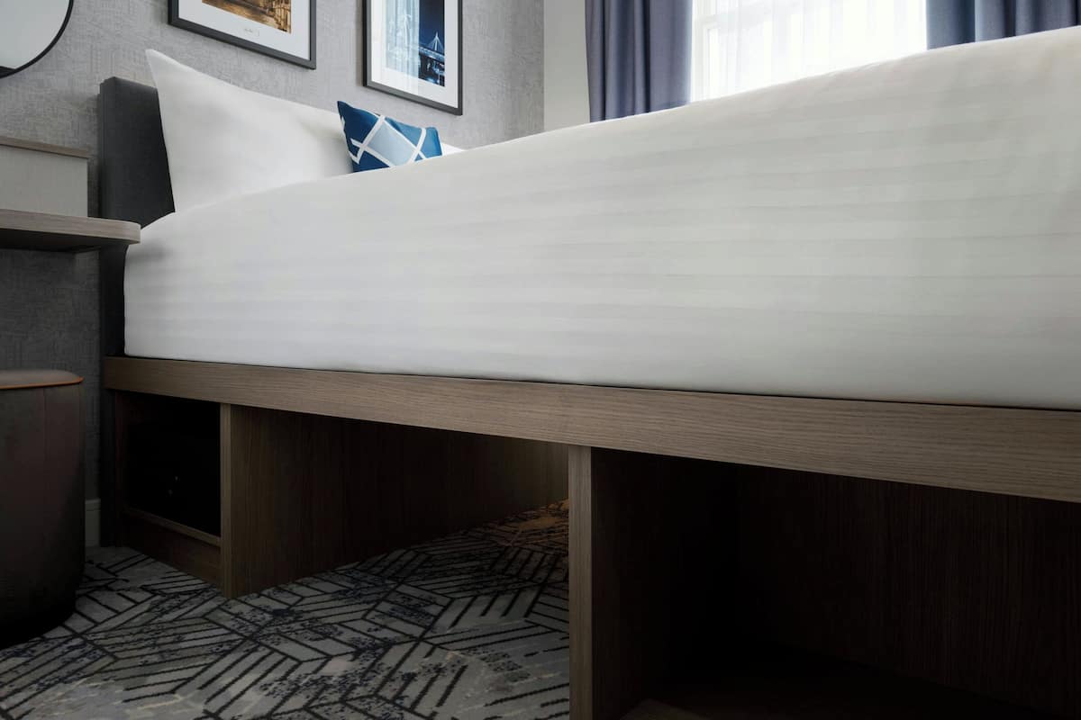 Uma cama bem arrumada com estrutura de madeira, uma mesa de cabeceira e um tapete estampado.