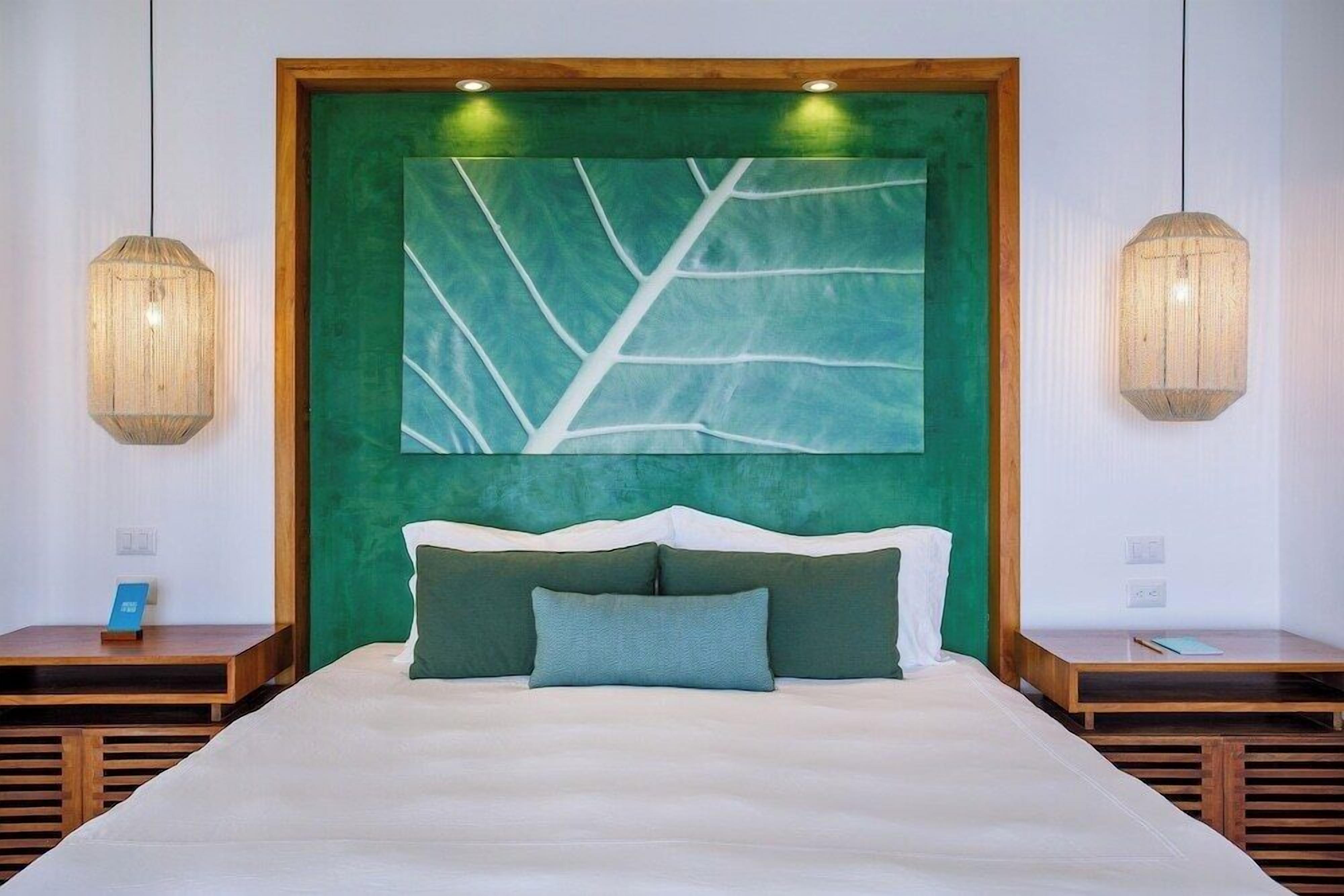 Uma cama com cabeceira estampada em folhagens verdes, duas mesas de cabeceira e duas luminárias pendentes.