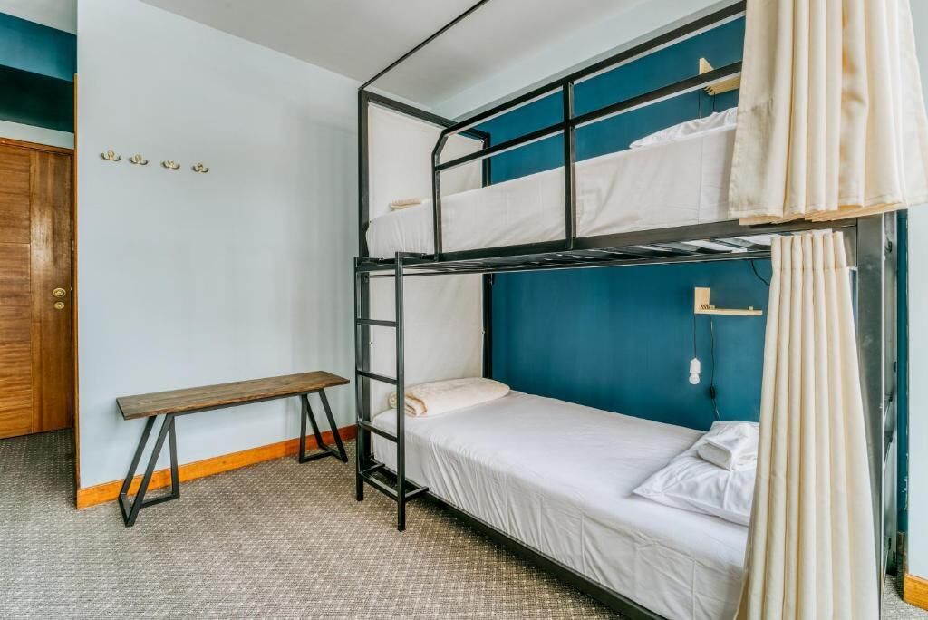Quarto com beliche, um banco de madeira, uma mesinha e cortina.