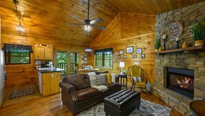 Interior acolhedor de uma cabana com teto de madeira, lareira, área de jantar e sala de estar com sofá e apoio para os pés.
