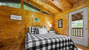 Um quarto de cabana de madeira acolhedor, com colcha xadrez, mesa de cabeceira com abajur e uma placa com a inscrição 'cada momento importa'.