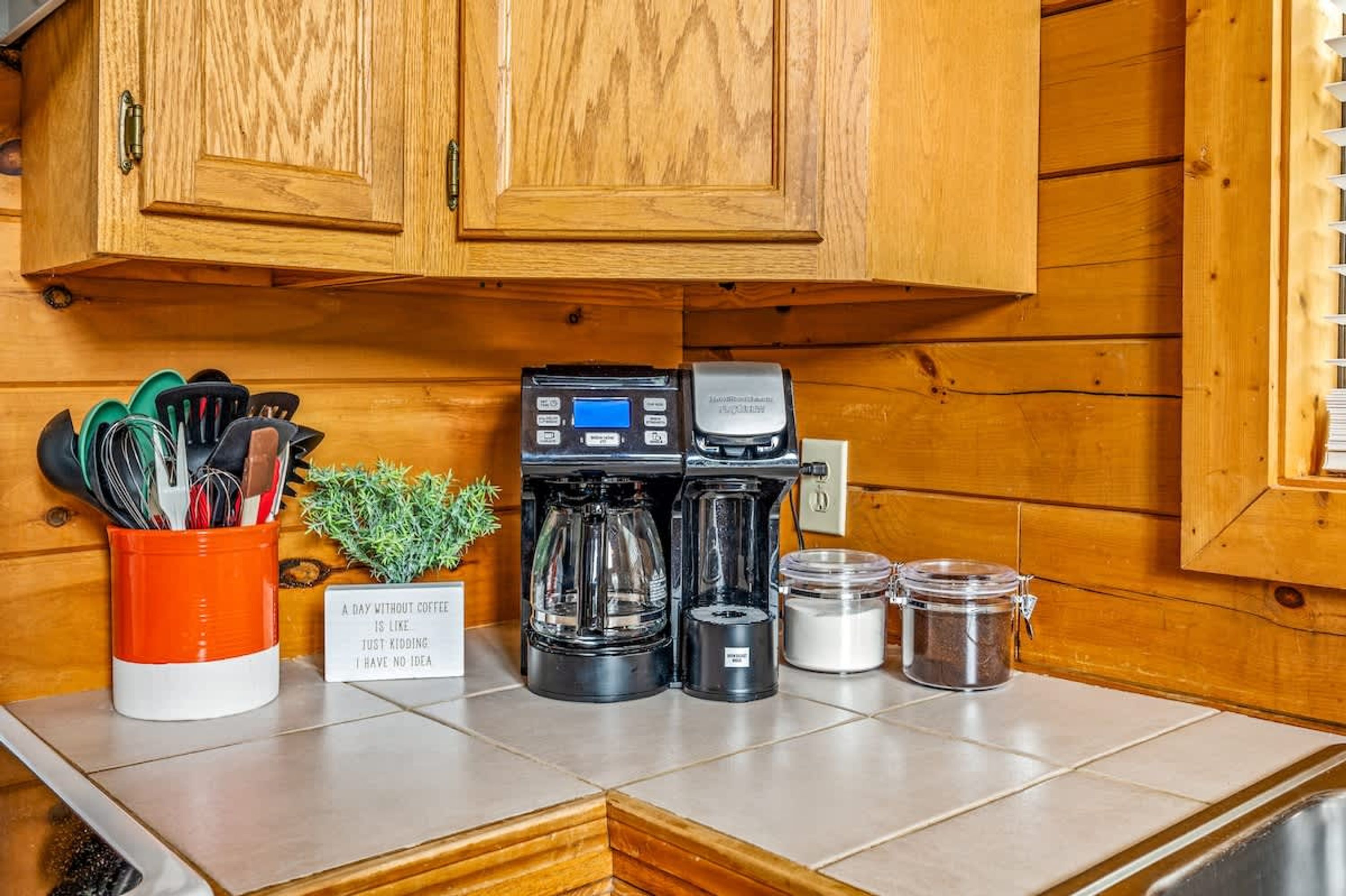 Uma cozinha com armários de madeira, cafeteira, liquidificador e uma placa com a inscrição 'Um dia sem café é como... brincadeira, não faço ideia'.
