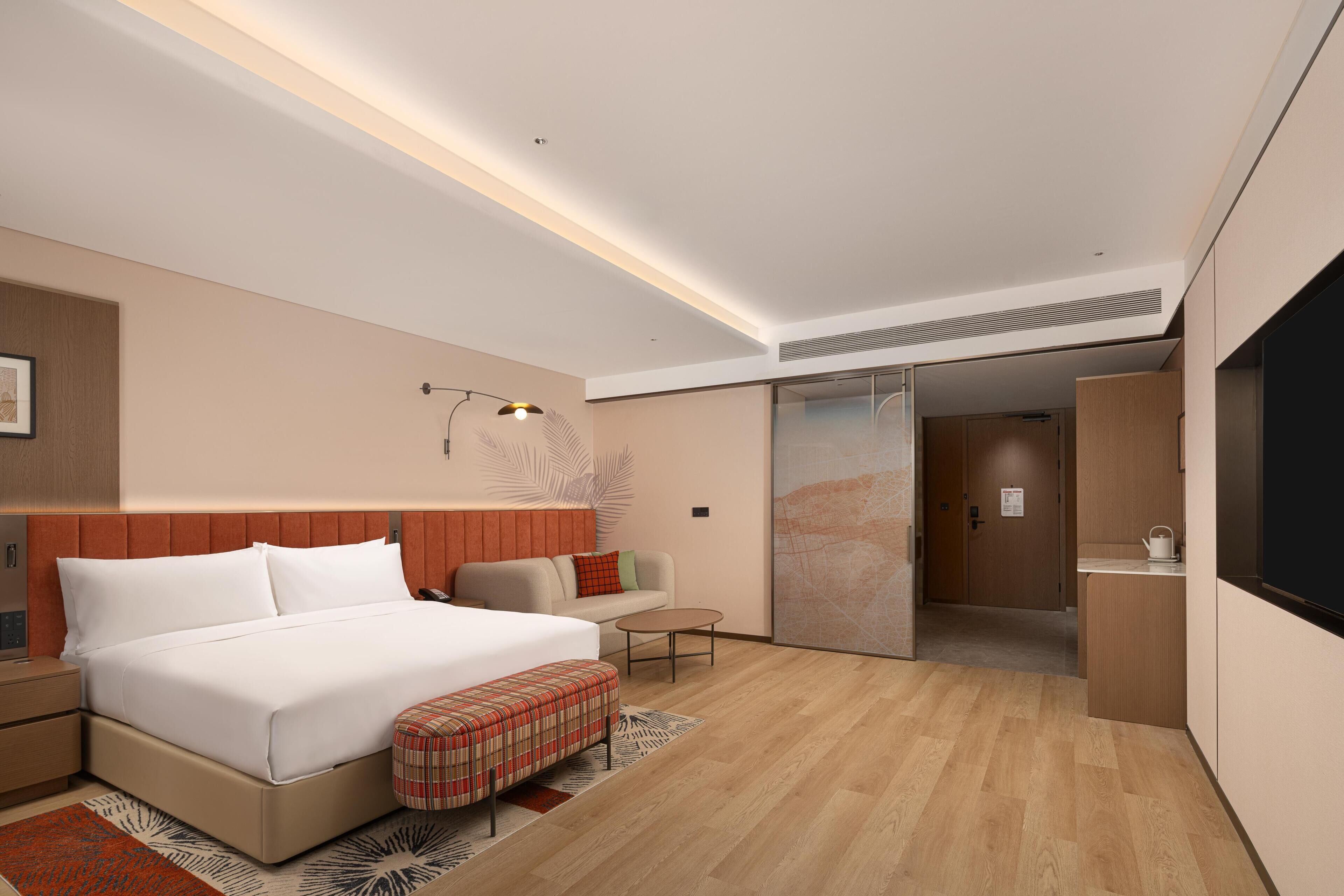 Quarto de hotel moderno com uma cama grande, um espaço de estar pequeno, uma televisão e um banheiro visível através de uma porta deslizante.