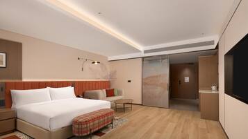 Quarto de hotel moderno com uma cama grande, um espaço de estar pequeno, uma televisão e um banheiro visível através de uma porta deslizante.
