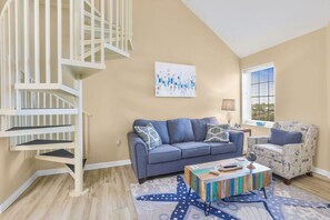 Uma sala de estar com um sofá azul, uma poltrona estampada, uma escada espiral branca e um quadro na parede.
