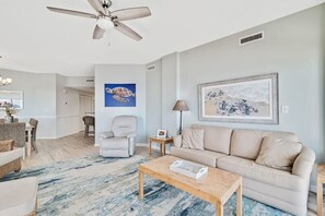Uma sala de estar com um sofá bege, uma mesa de centro, um ventilador de teto, um quadro e uma área de jantar com mesa e cadeiras.