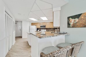 Uma cozinha moderna com armários brancos, bancada de granito e micro-ondas. Há uma cadeira de vime e uma pequena ilha com banquetas.