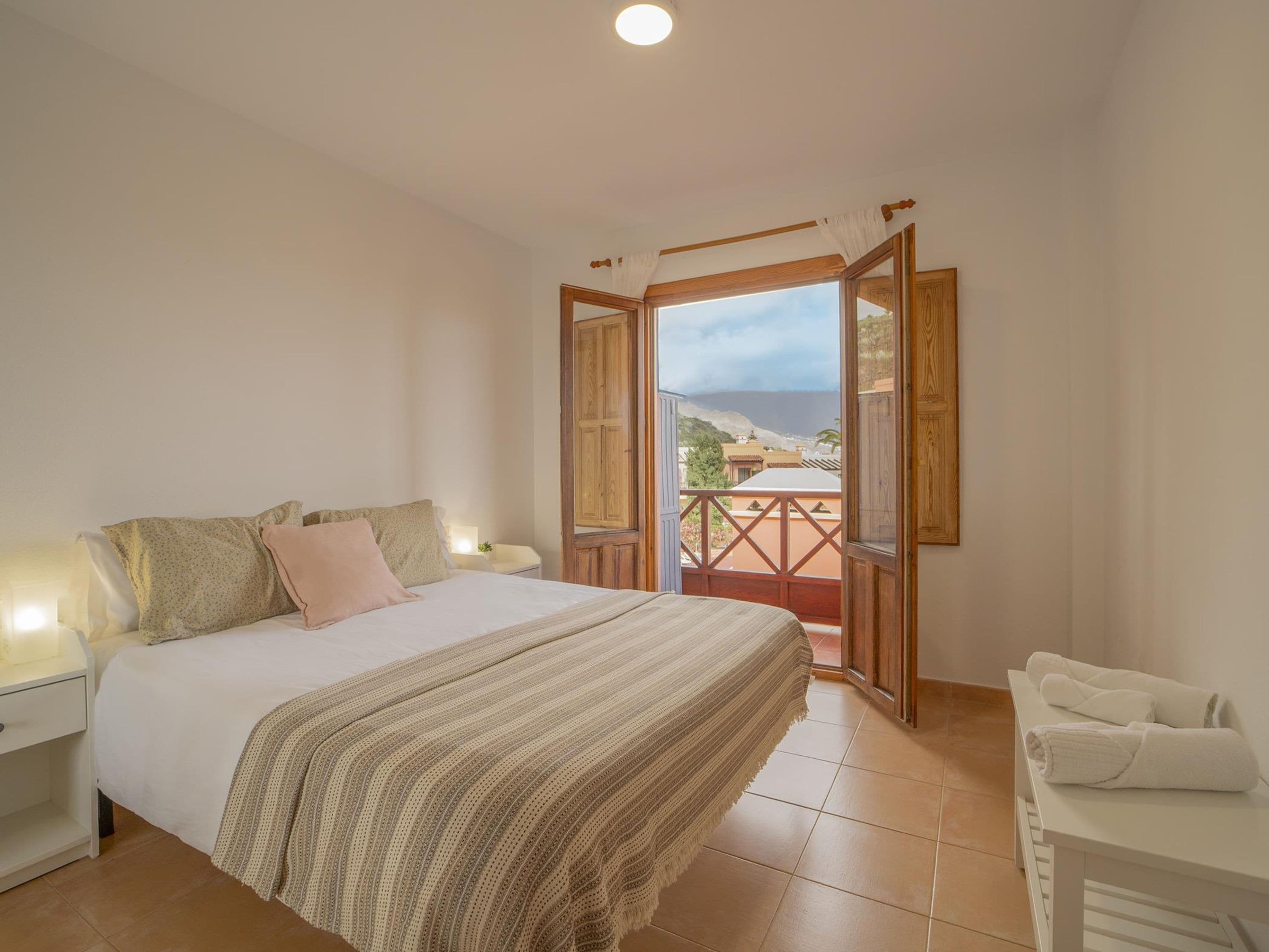 Quarto com cama, mesa de cabeceira e vista para o exterior através de uma porta aberta.