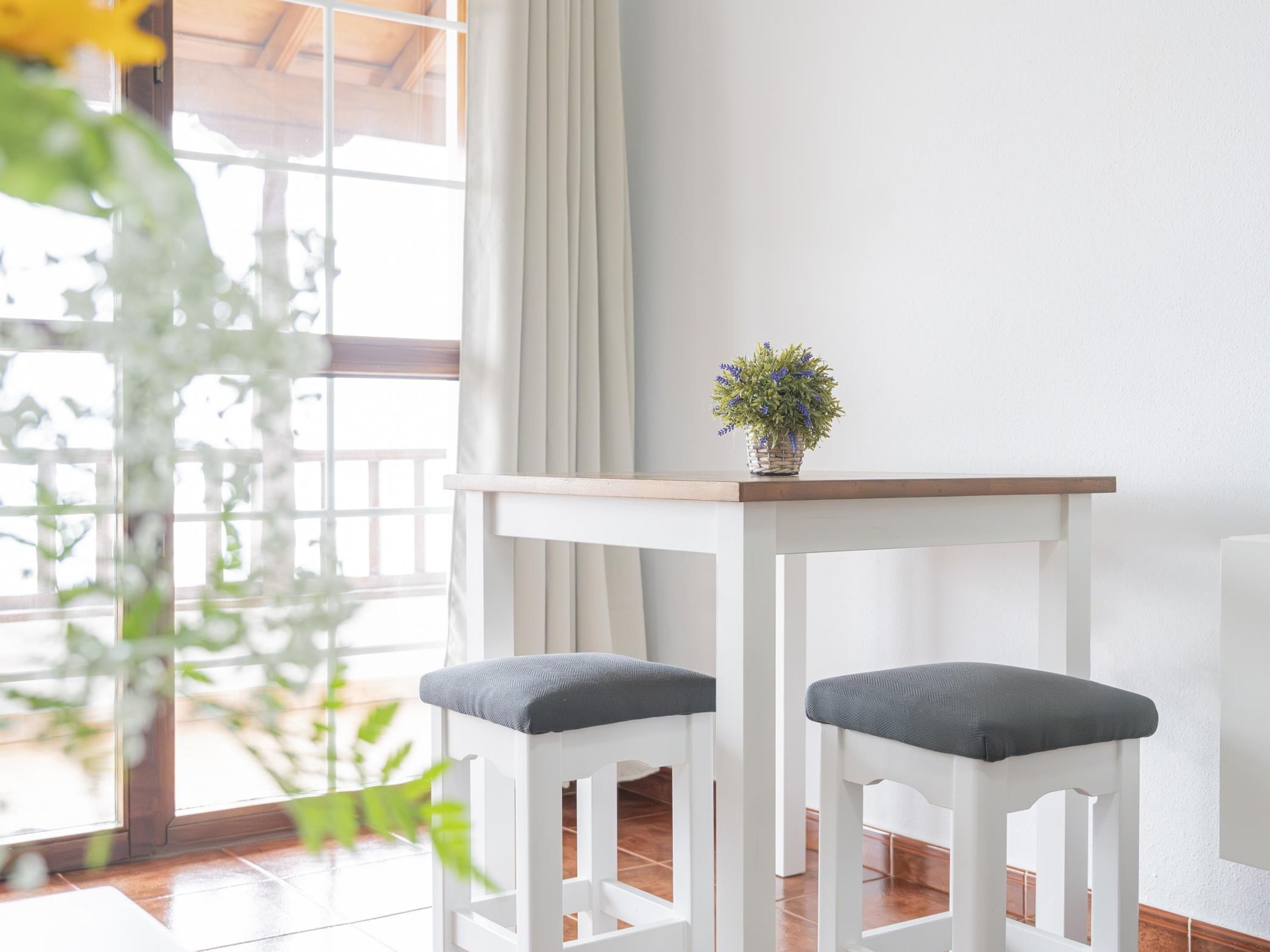 Uma pequena mesa branca com uma cesta de palha contendo uma planta, dois banquinhos com almofadas cinzas e uma parede branca.