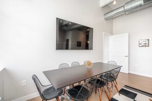 Uma área de refeições moderna com mesa de madeira escura e cadeiras de estrutura de arame preto, um espelho grande na parede e uma porta branca com uma pequena imagem acima.