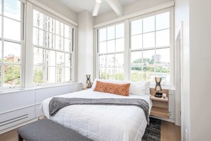 Quarto com cama grande, duas mesas de cabeceira com abajures, um banco e vista para edifícios através de janelas amplas.