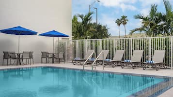 Piscina com espreguiçadeiras e guarda-sóis, cercada por um muro branco e palmeiras.