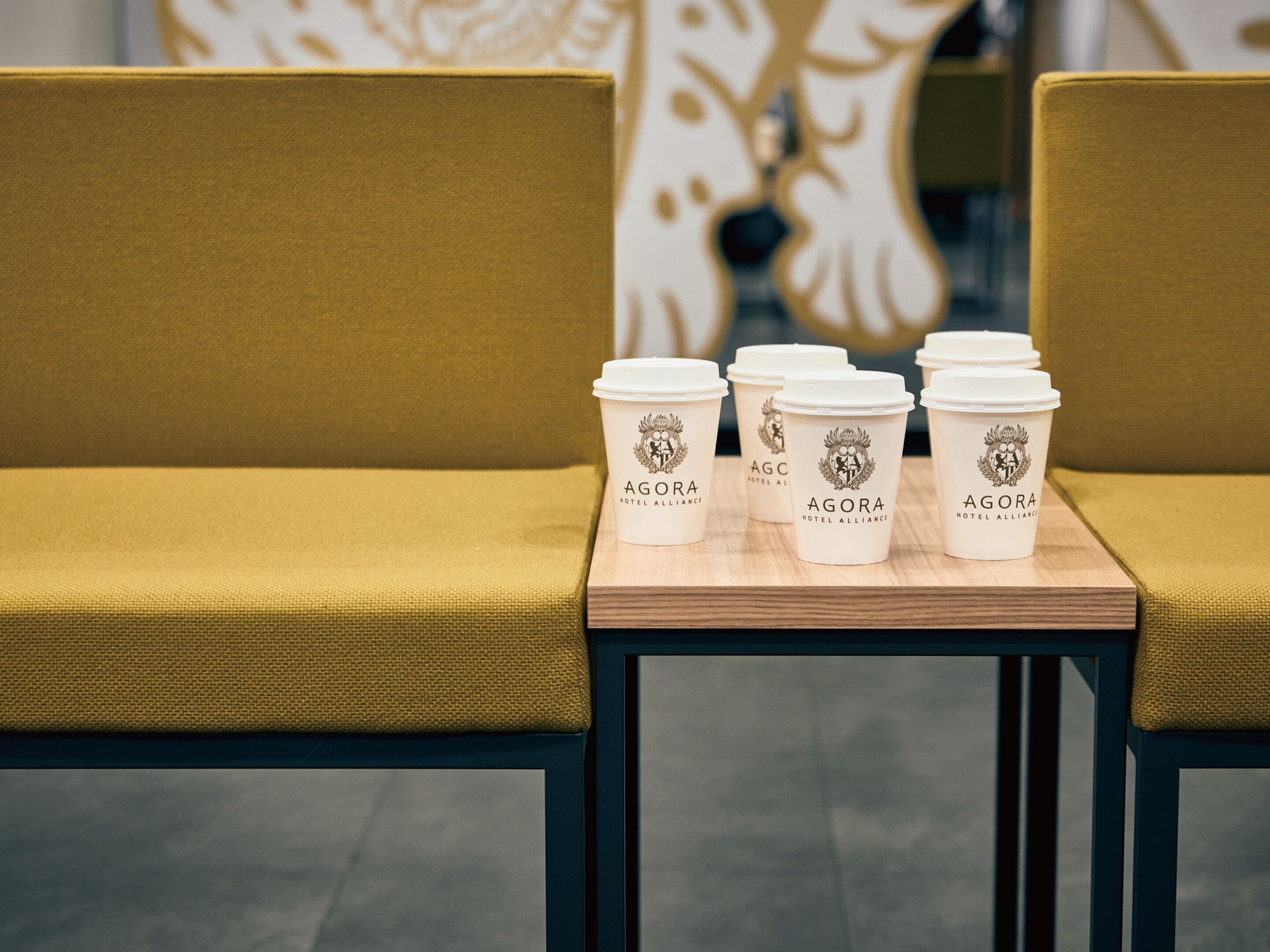 Uma mesa com seis xícaras de café identificadas como 'Agora Hotel Alliance' entre duas cadeiras amarelas.