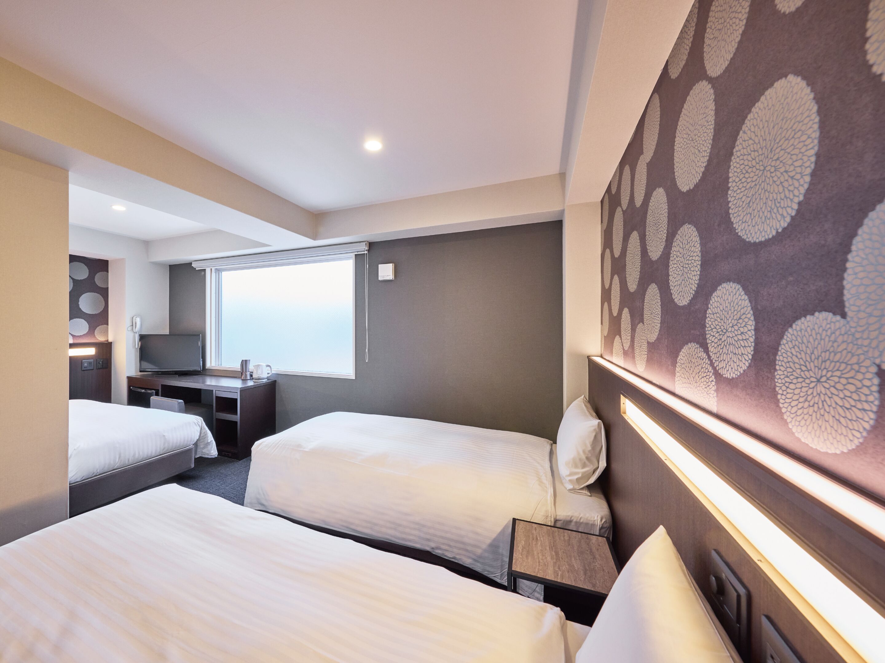 Quarto de hotel com três camas, uma escrivaninha com televisão, uma janela grande e parede com padrão.