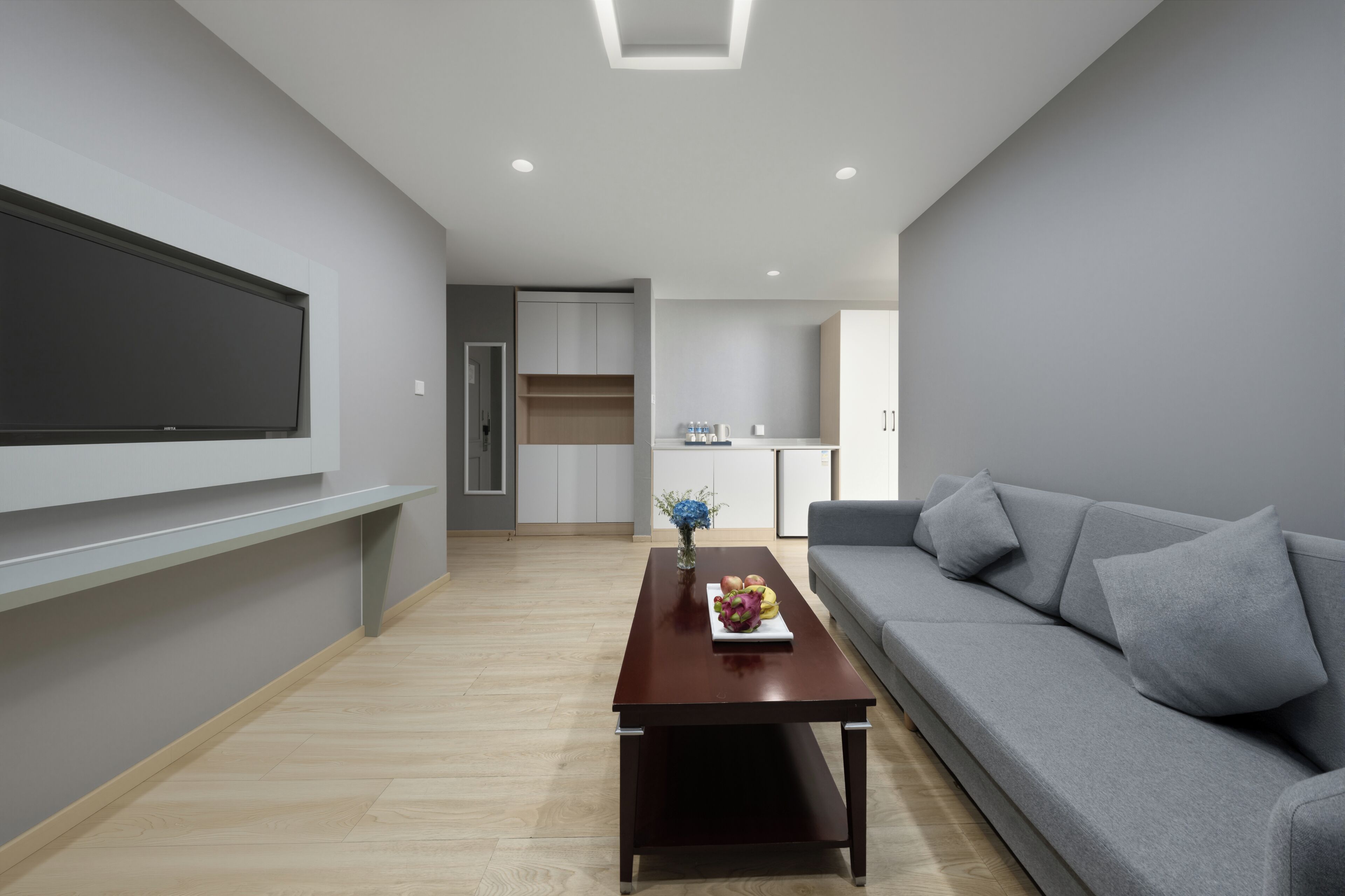 Uma sala de estar moderna com uma televisão de tela plana, um sofá cinza, uma mesa de centro de madeira e uma cozinha ao fundo.