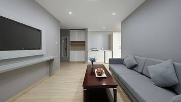 Uma sala de estar moderna com uma televisão de tela plana, um sofá cinza, uma mesa de centro de madeira e uma cozinha ao fundo.