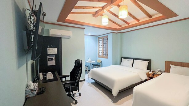 Quarto de hotel com cama, escrivaninha e cadeira, televisão e teto com vigas de madeira e luminárias pendentes.