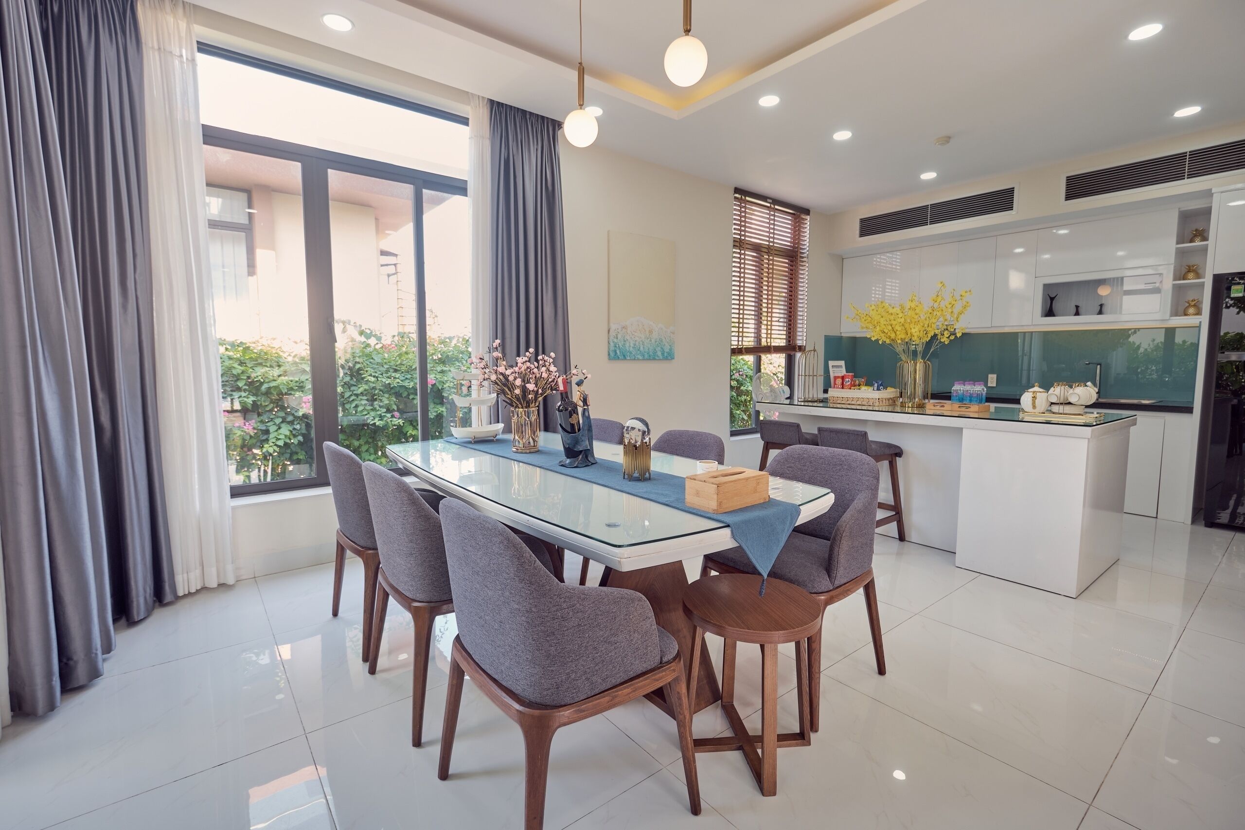 Uma área de jantar moderna com mesa de vidro, cadeiras estofadas em cinza e um vaso central com flores.
