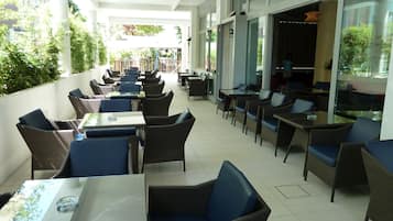 Área de café ao ar livre com móveis de vime e mesas de vidro, cercada por paredes de vidro e vista para o verde.