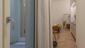 Quarto de hotel com piso de madeira, cama com cobertura verde, uma cadeira, uma mesinha e uma cozinha compacta.