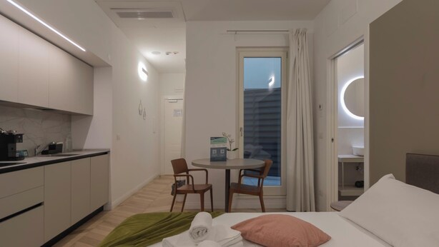Quarto de hotel com cama, pequena cozinha, área de refeições e vista do céu pela janela.