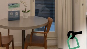 Quarto com uma mesa redonda, duas cadeiras, um vaso de planta, um aviso na parede e uma janela com cortinas.