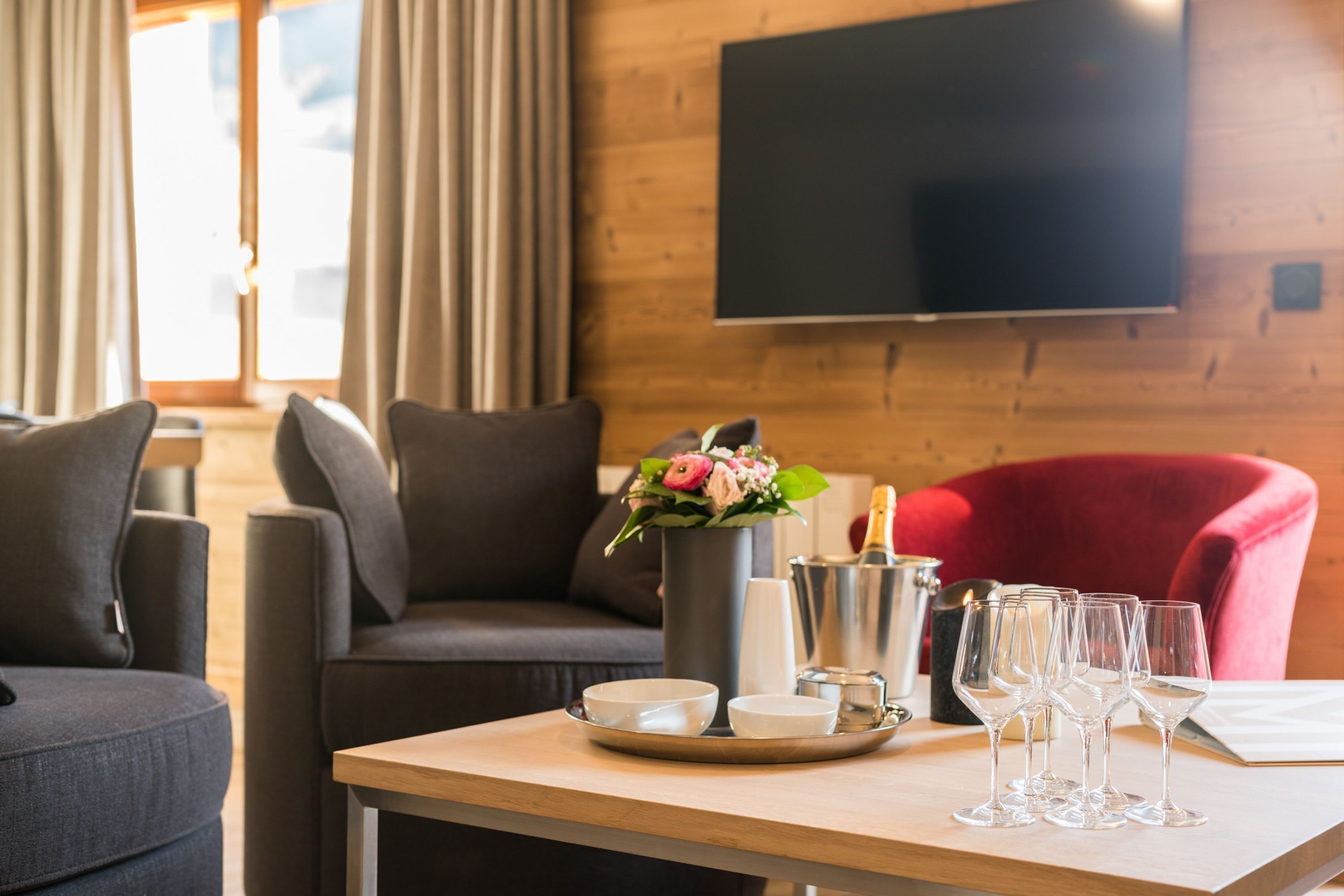 Quarto de hotel com parede de madeira, TV de tela plana, poltrona vermelha, sofá cinza, mesa de madeira com bandeja de taças de vinho, um vaso com flores e uma garrafa de champanhe.