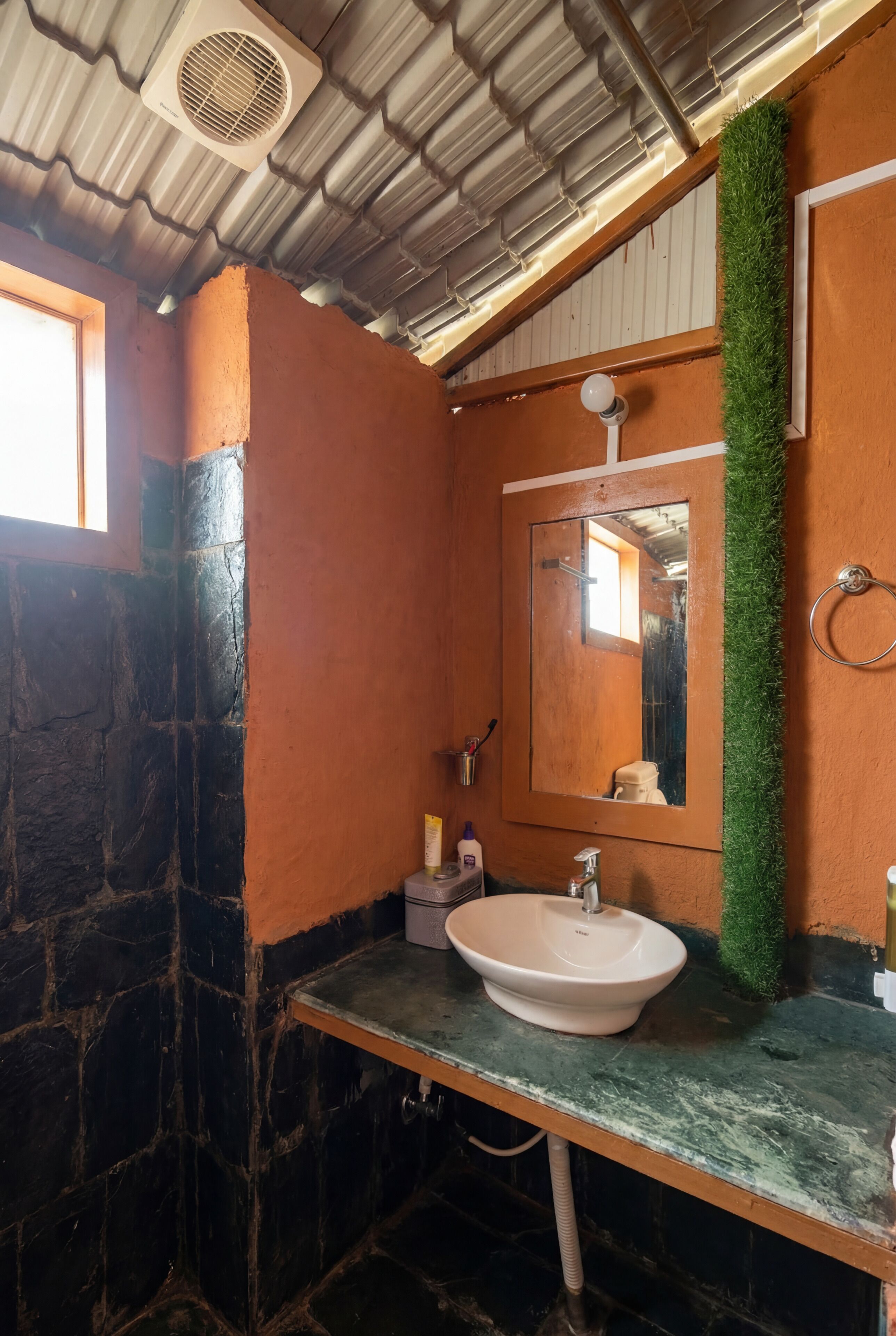 Casa de banho com parede de pedra, bancada verde, lavatório branco, espelho e ventoinha no teto.