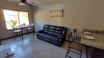 Uma sala de estar com sofá de couro preto, mesa de jantar com cadeiras, ventilador de teto e um quadro na parede.