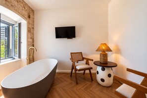 Uma casa de banho moderna com uma banheira independente, uma cadeira de madeira, uma mesa redonda com uma lâmpada e uma televisão na parede.