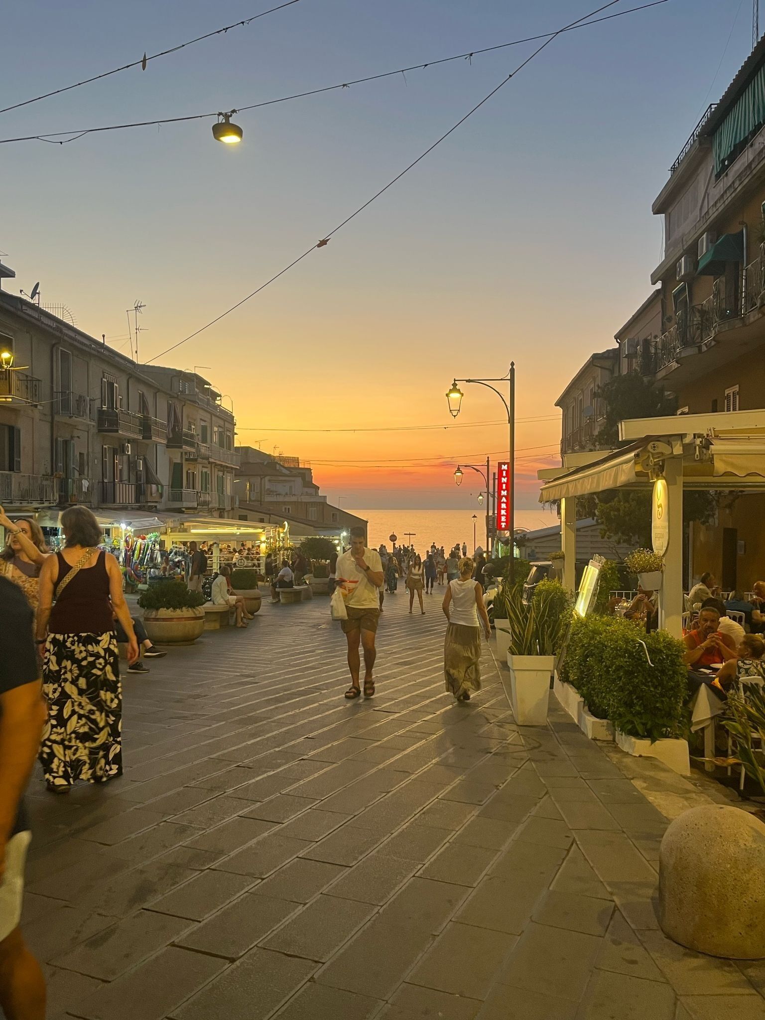 Cena de rua movimentada ao pôr do sol, com pessoas caminhando, postes de luz e edifícios.