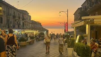 Cena de rua movimentada ao pôr do sol, com pessoas caminhando, postes de luz e edifícios.