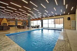 Piscina interna com chão ladrilhado, teto de madeira com iluminação linear e parede de tijolos com lareira.