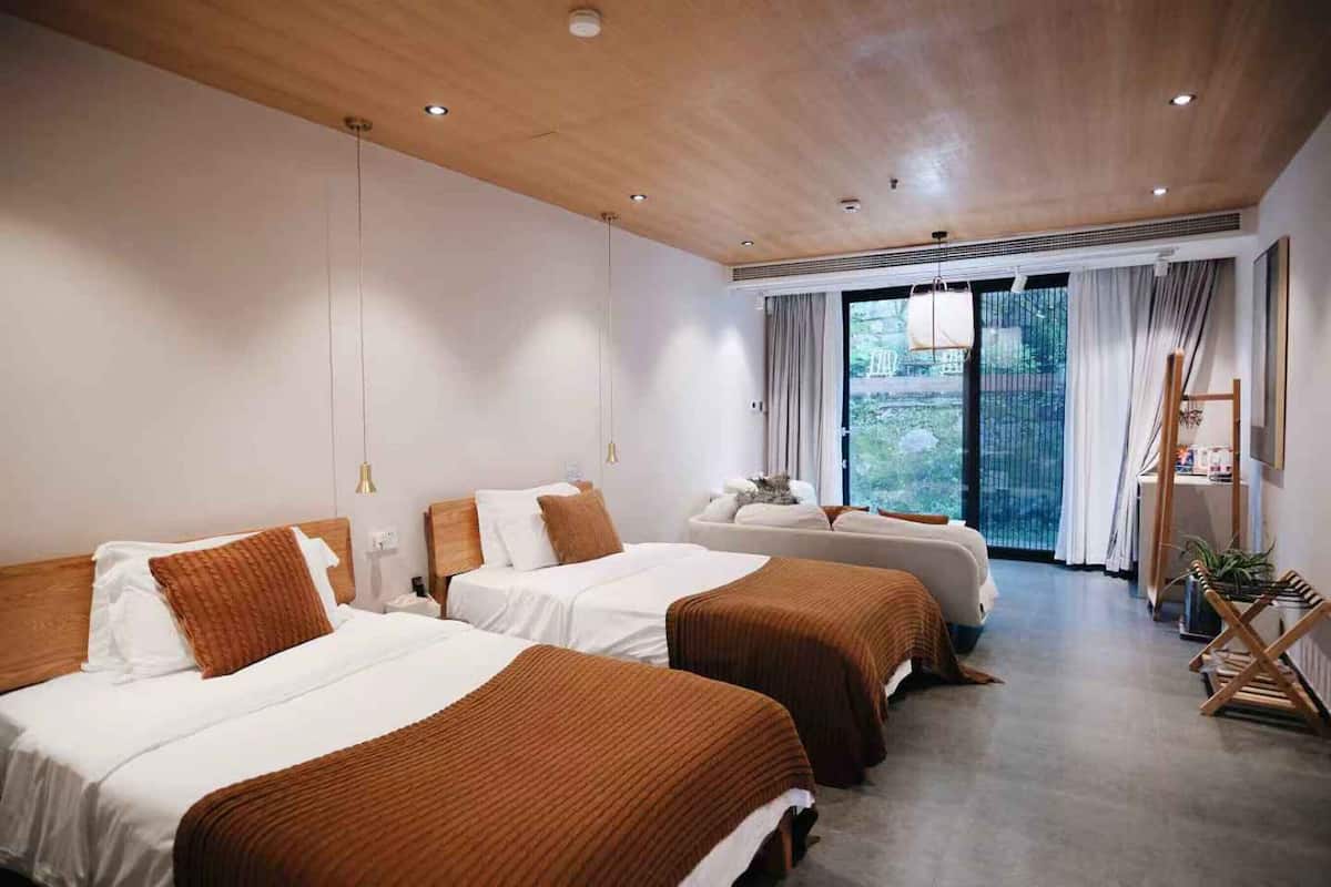 Quarto de hotel com três camas, um sofá, uma escrivaninha e uma janela grande com cortinas.