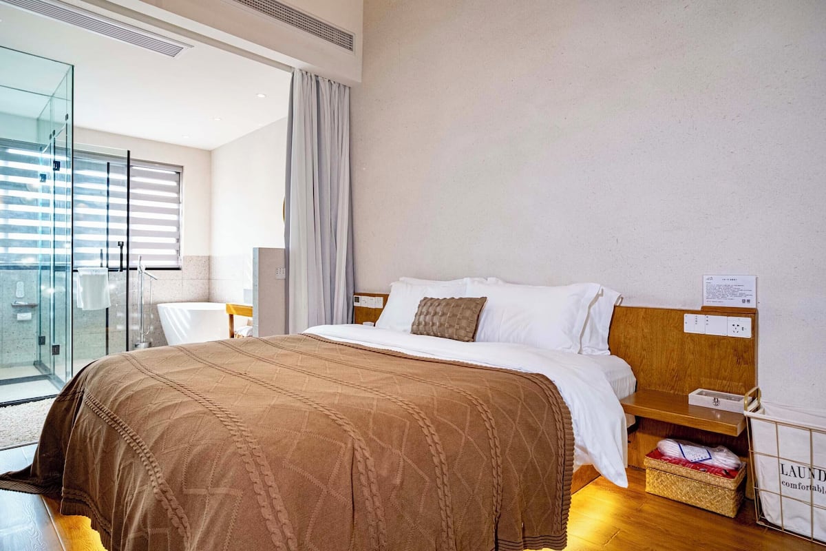 Quarto de hotel com uma cama grande, mesa de cabeceira, box de vidro e vista para o exterior.