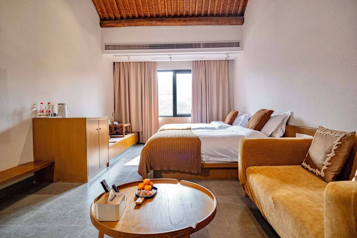Quarto de hotel com cama, sofá, mesa de centro de madeira com frutas, escrivaninha com computador e janela com cortinas.